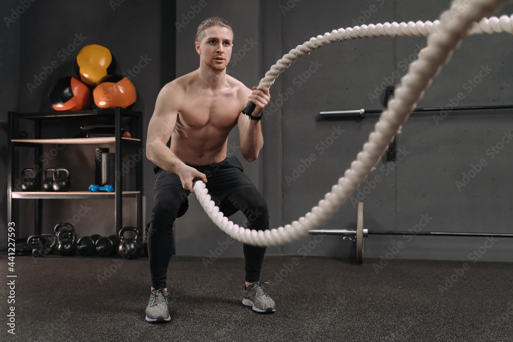 Muscular handsome athletic guy working out with battle rope at dark gym ...