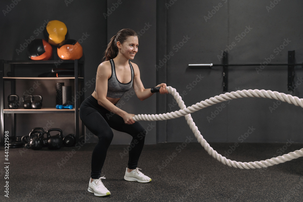 Muscular young smiling woman working out with battle ropes at dark gym ...