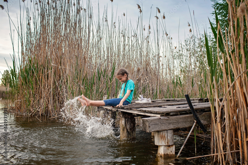 Cute preteen girl sits on a wooden pier by the lake and stirs feet in ...