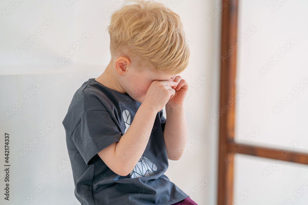 little boy with blond hair, rubs his eyes and cries with his back ...