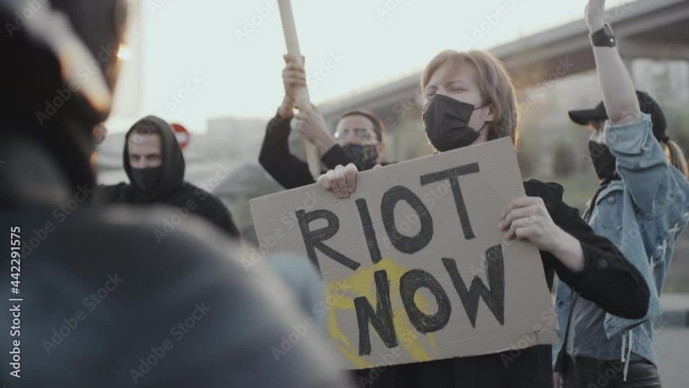 Handheld slowmo shot of angry masked people with signs chanting and ...