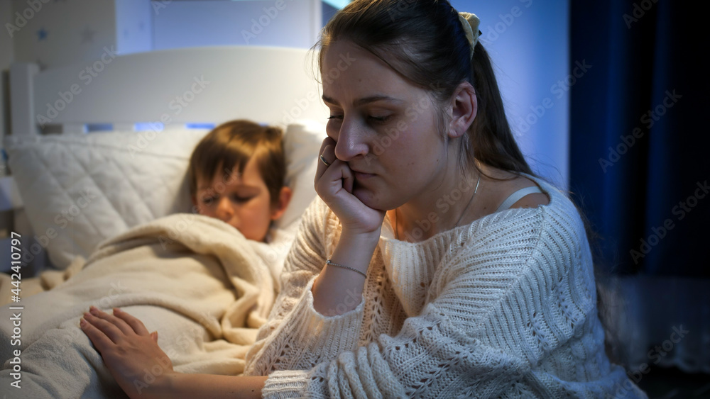 Upset, sad and stressed mother sitting next to bed of her sleeping ...