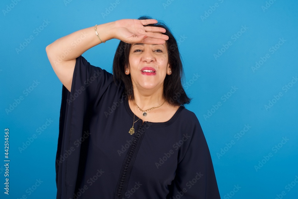 middle aged Arab woman standing against blue background wiping forehead ...
