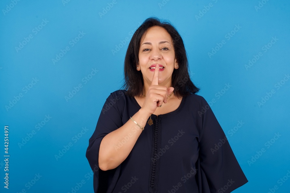 Smiling middle aged Arab woman standing against blue background makes ...