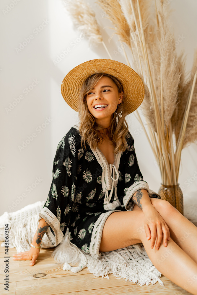 Laughing blond woman in straw hat and boho dress posing in studio over ...