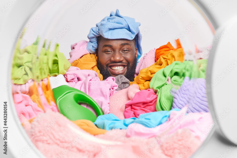 Cheerful dark skinned man covered with pile of laundry has fun poses ...