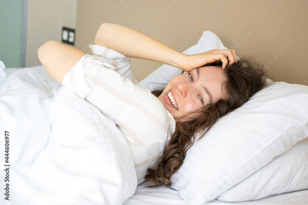 Pretty brunette young woman waking up lying in white comfortable bed, stretching arms raising ...