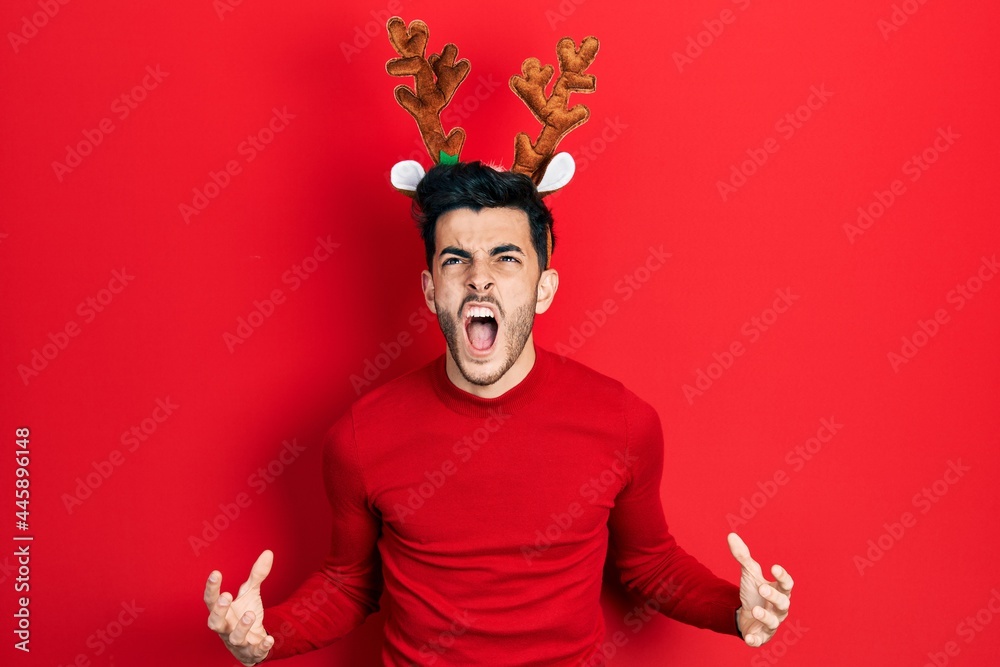 Young hispanic man wearing cute christmas reindeer horns crazy and mad ...