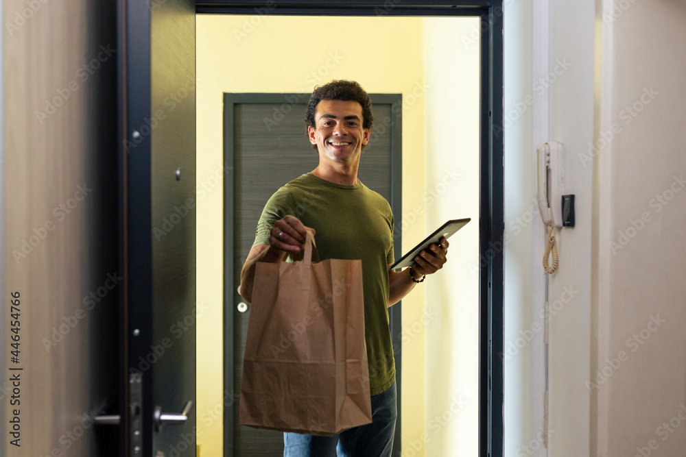 Smiling delivery man in front of the door with tablet and paper bag ...