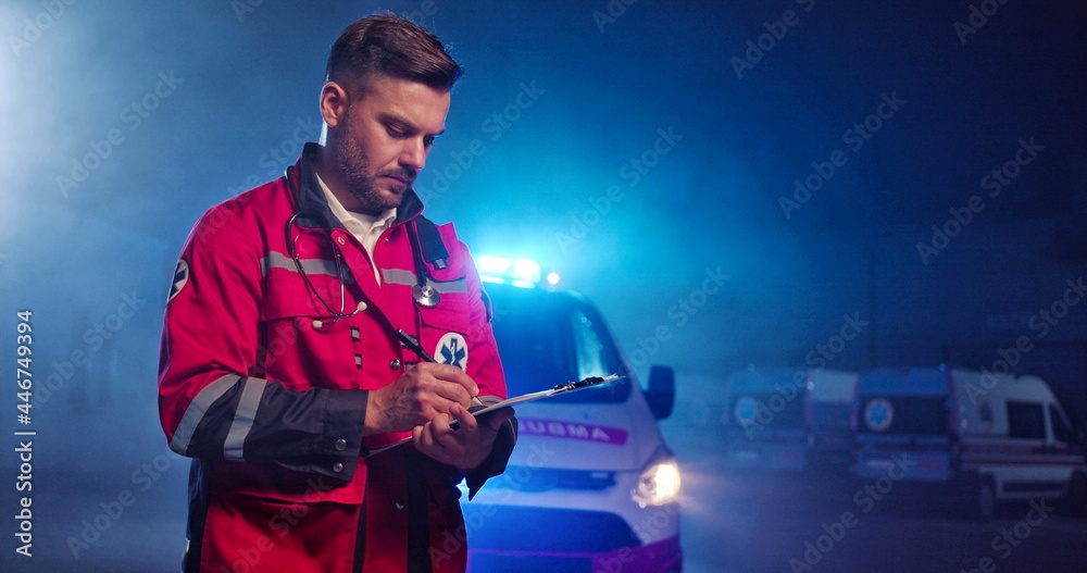 Handsome young Caucasian male paramedic in red uniform writing with pen ...
