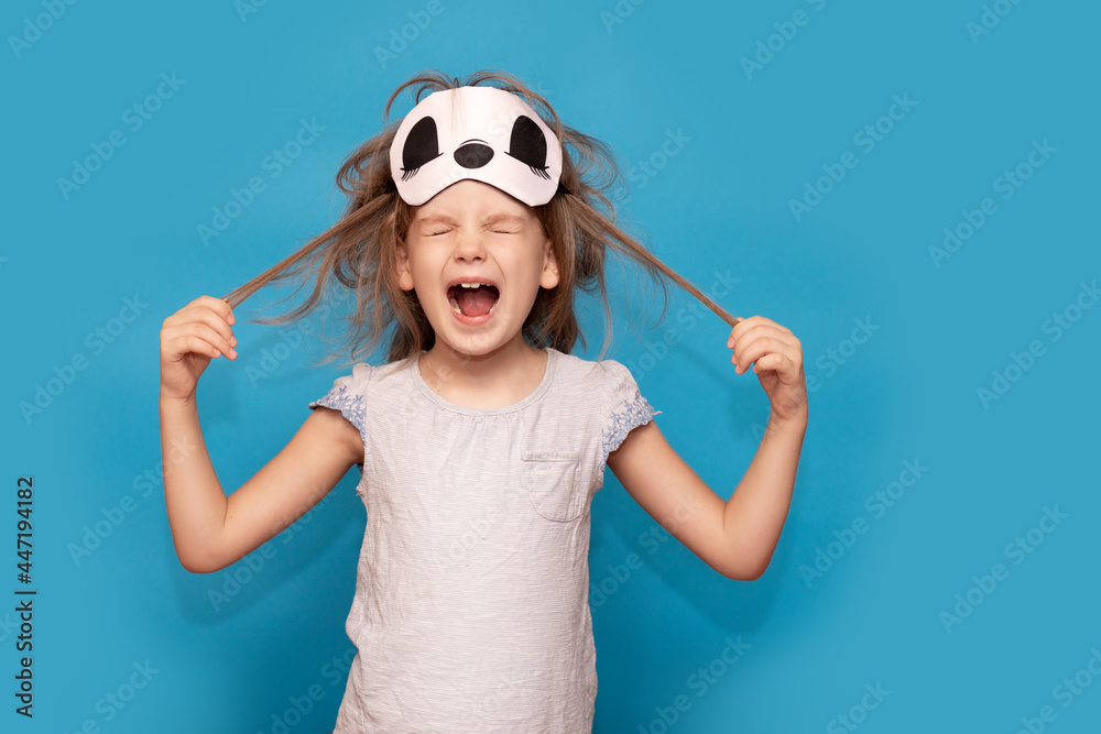 Spoiled little girl in sleeping mask, tearing her hair isolated on blue ...