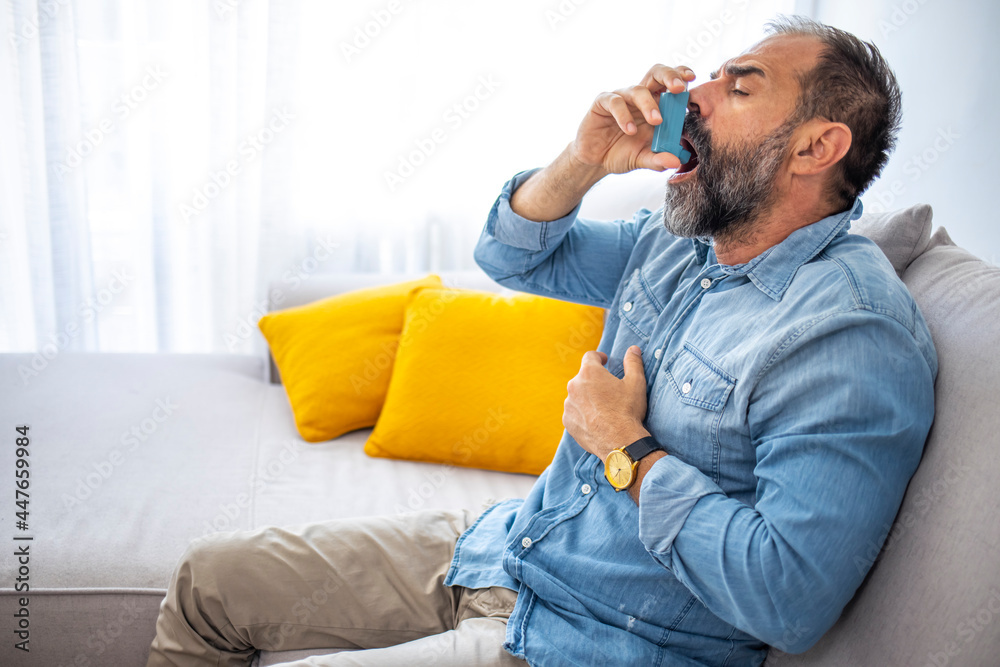 Side view of an asthmatic man using an inhaler sitting on a couch in ...
