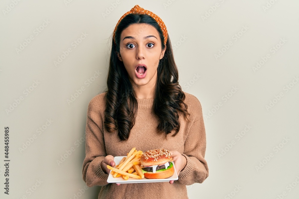 Young hispanic woman eating a tasty classic burger afraid and shocked ...