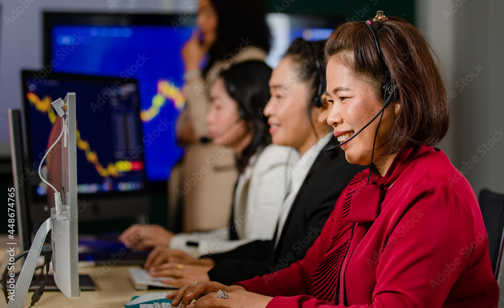 Group of Asian female customer service operators wears microphone headsets sit smiling in front ...