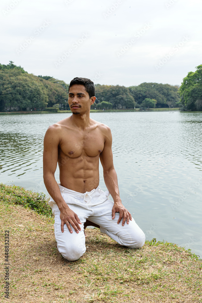 Foto de Brazilian young man sitting on his knees by a lake, shirtless ...