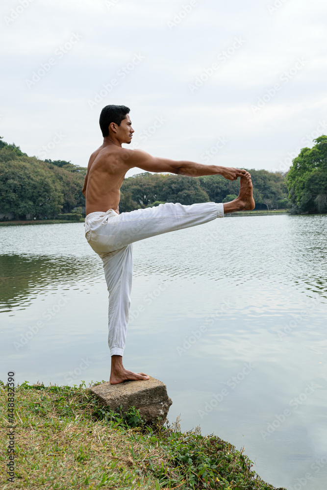 Young Brazilian man standing beside a lake, shirtless, stretching one ...