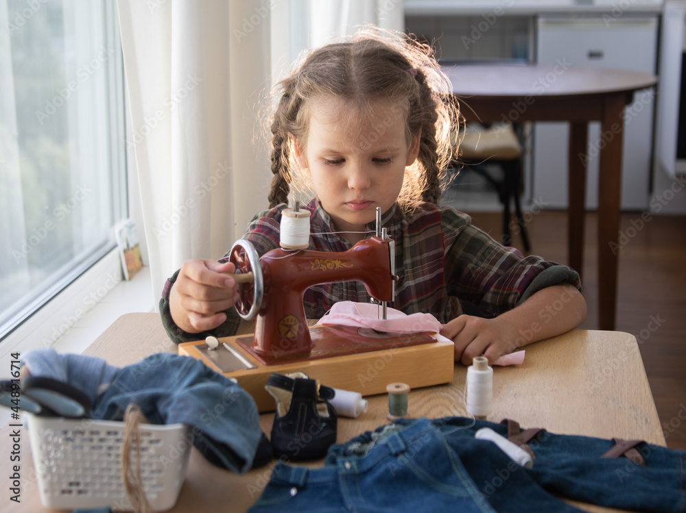 A little girl sews clothes for dolls on a toy sewing machine, a child ...