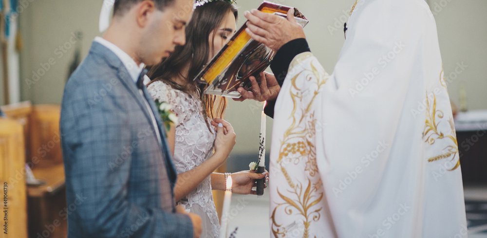 Stylish bride and groom kissing holy bible from priest hands during ...