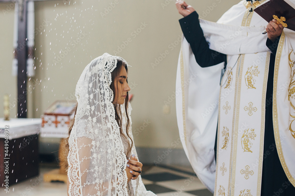 Priest blessing with holy water stylish bride in kerchief at altar ...