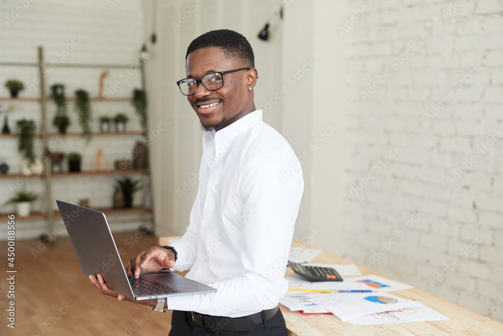 Smiling African man working in a modern office behind a laptop and ...