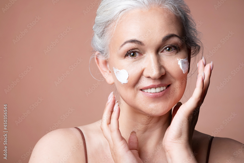 Happy mature gray-haired female moisturizing skin by facial cream ...