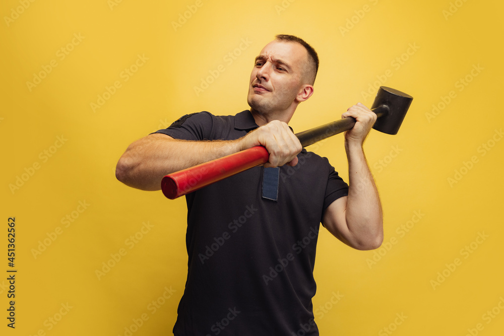 One young brutal man, firefighterposing with hammer over yellow studio ...