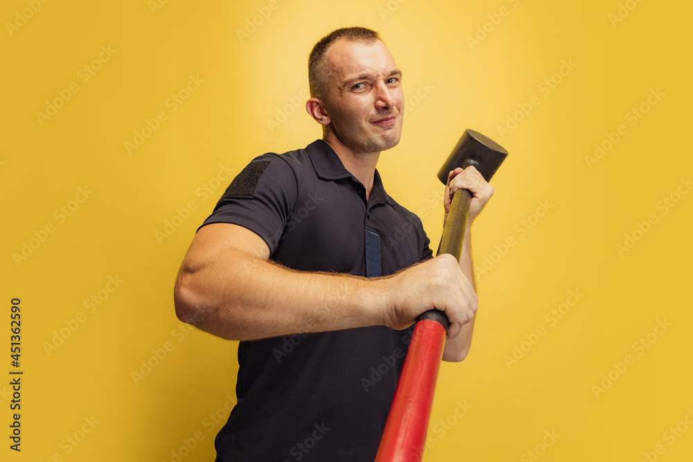 One young brutal man, firefighterposing with hammer over yellow studio ...