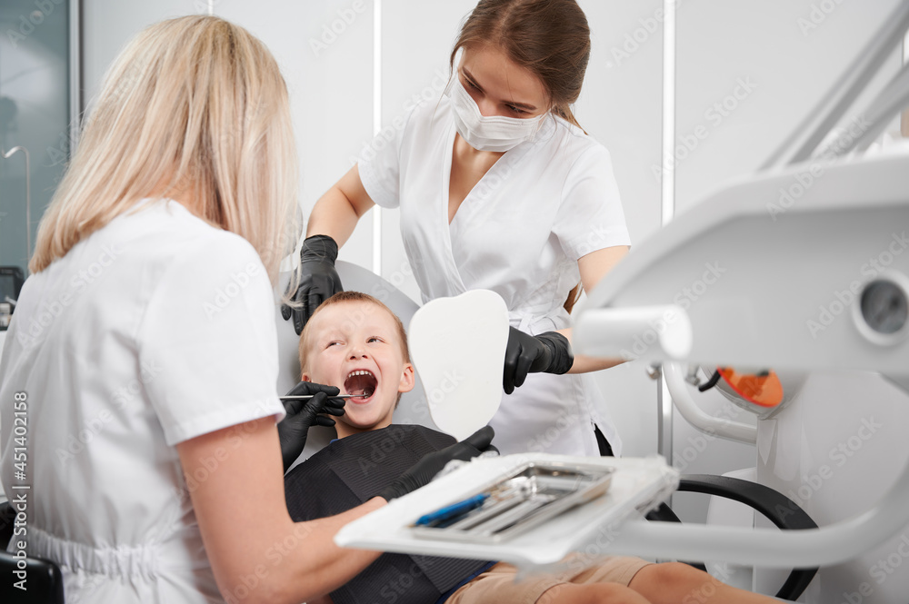 Adorable little boy sitting in dental chair while two female dentists checking kid teeth ...