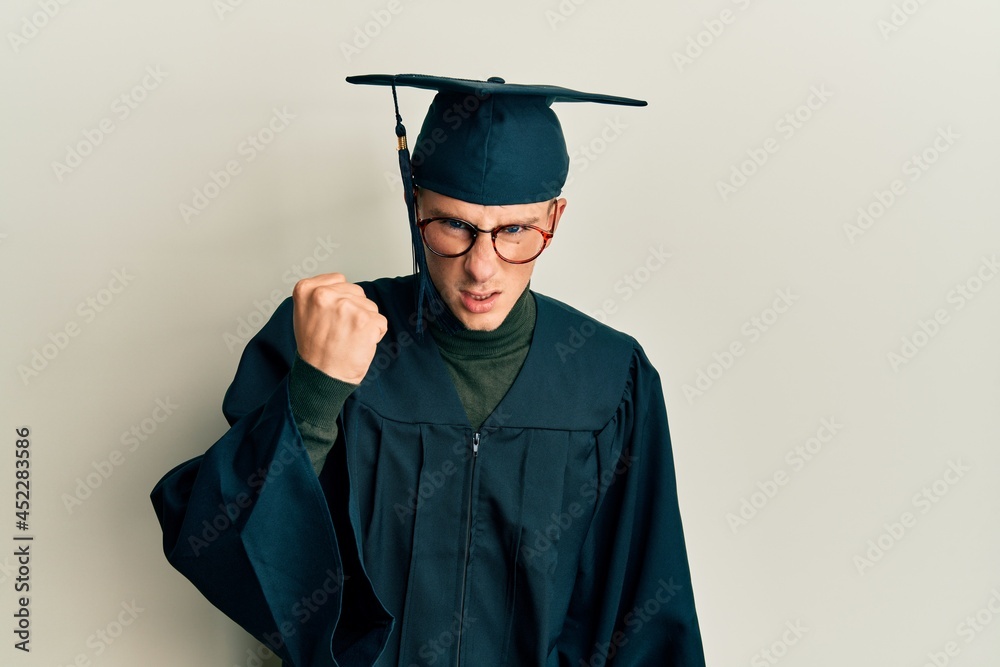 Young caucasian man wearing graduation cap and ceremony robe angry and ...