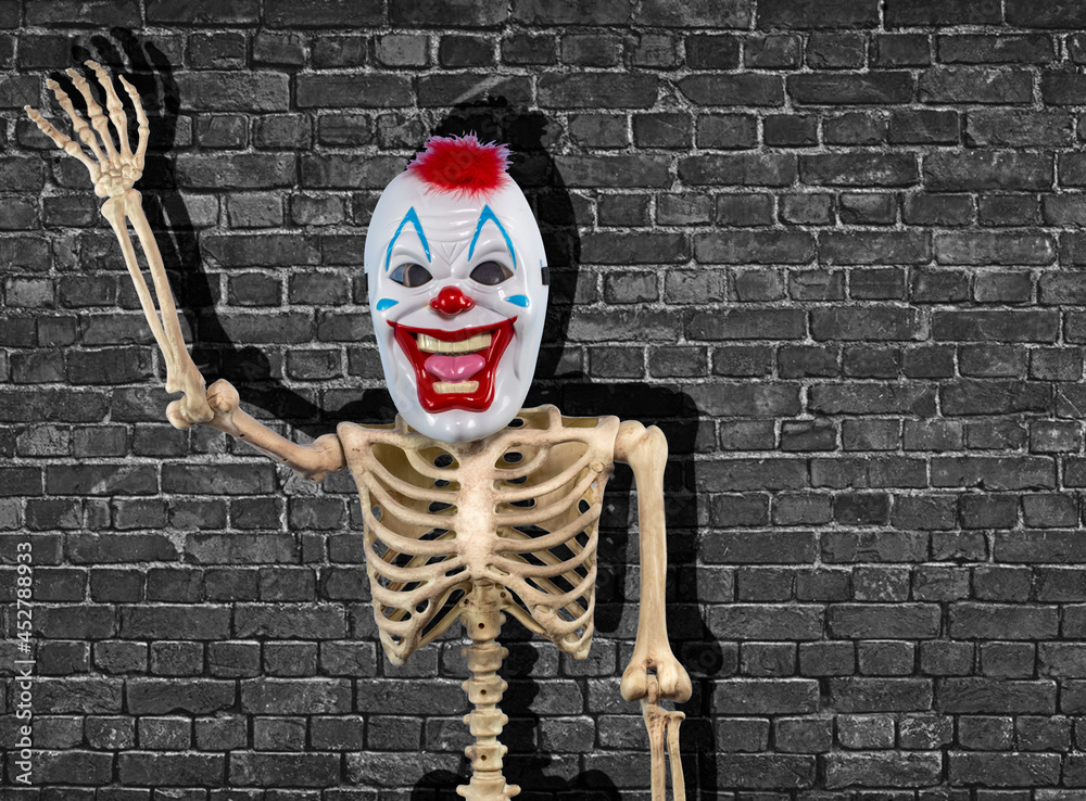 Skeleton wearing a clown Halloween mask in front of a black brick wall ...
