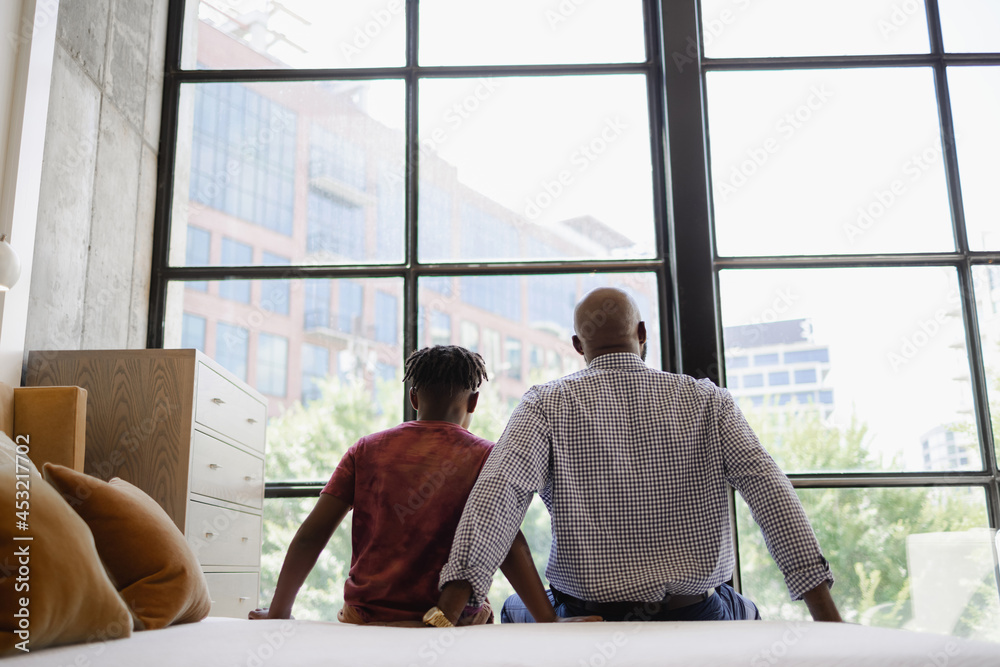 Rear view of Black father and son sitting on bed in front of window ...