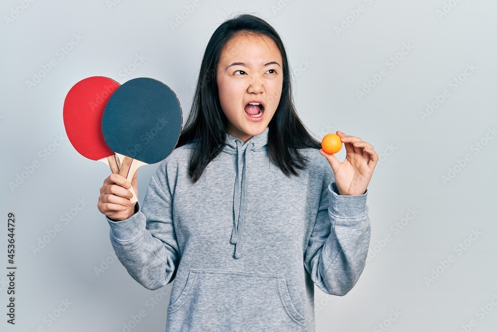 Young chinese girl holding red ping pong rackets and ball angry and mad ...