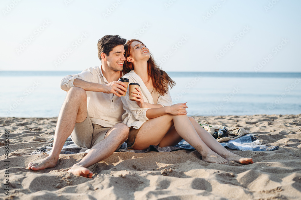 Full body young happy smiling couple family two man woman in white clothes hug sit on sand plaid ...