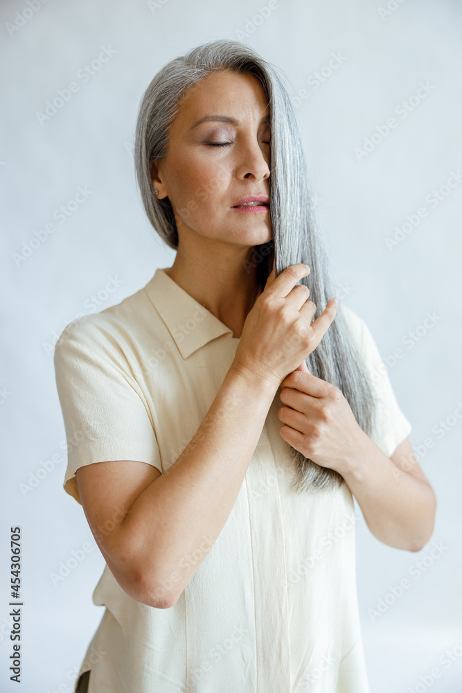Languid middle aged Asian woman in blouse strokes lock of silver hair ...