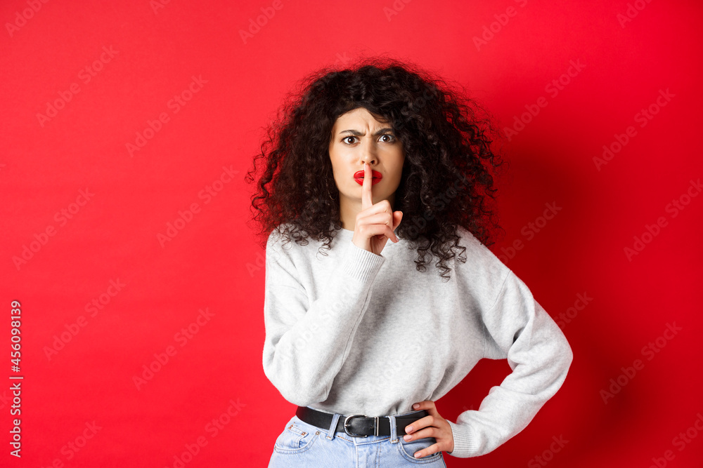 Angry young woman with curly hair and red lips, frowning and shushing ...