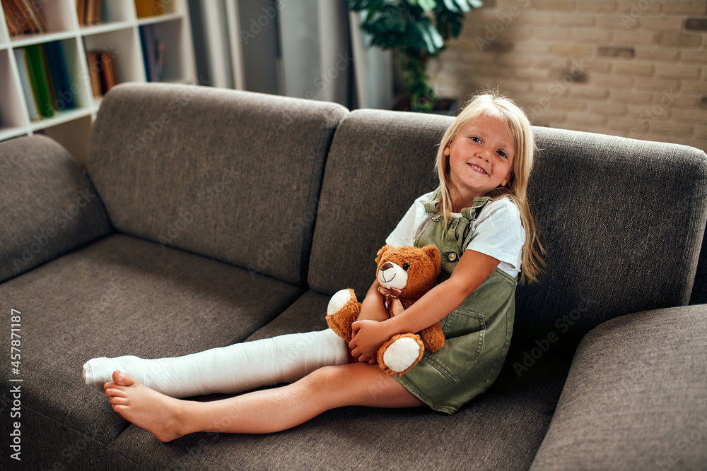 A cute little girl with a broken leg in a cast, sits hugging a teddy ...