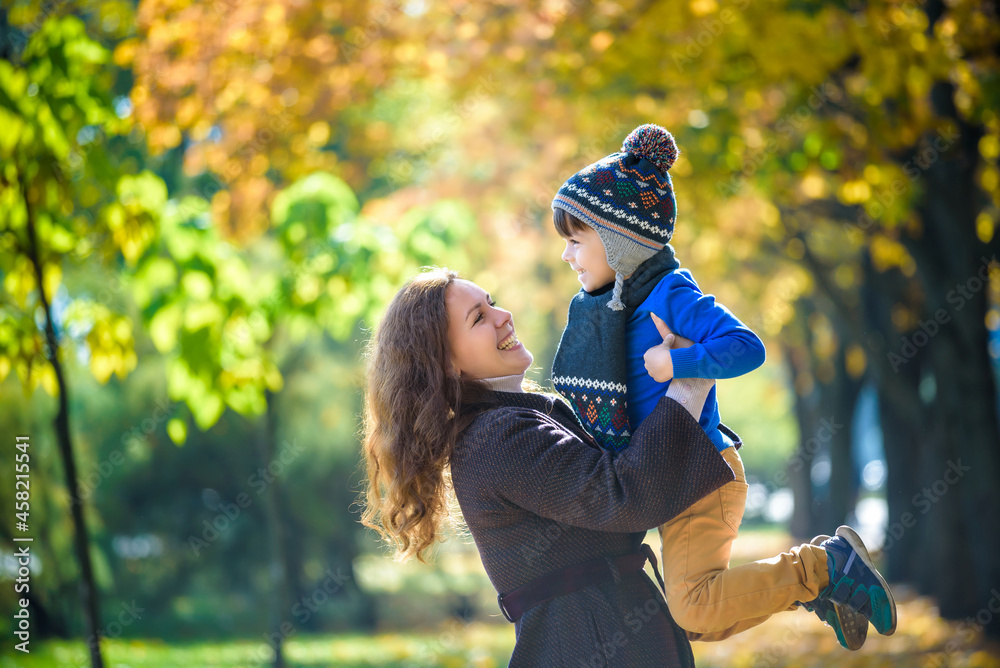 Cute, happy, boy smiling and hugging with his mom among yellow leaves ...