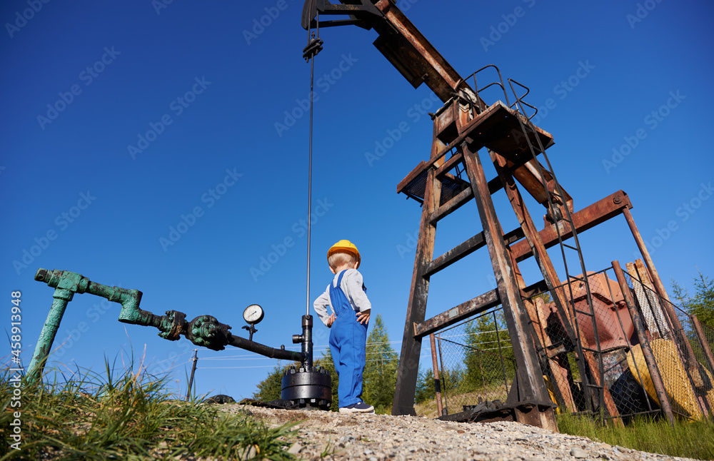 Cute child in work overalls standing near petroleum pump jack under ...