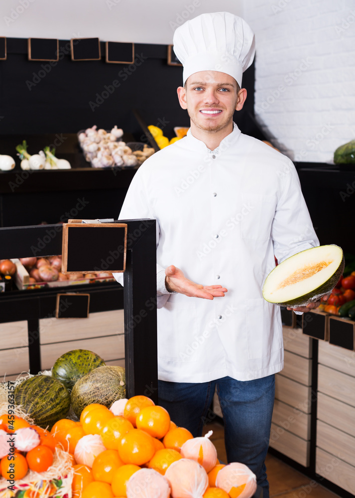 Young smiling male cook in white uniform choosing fruits in grocery ...