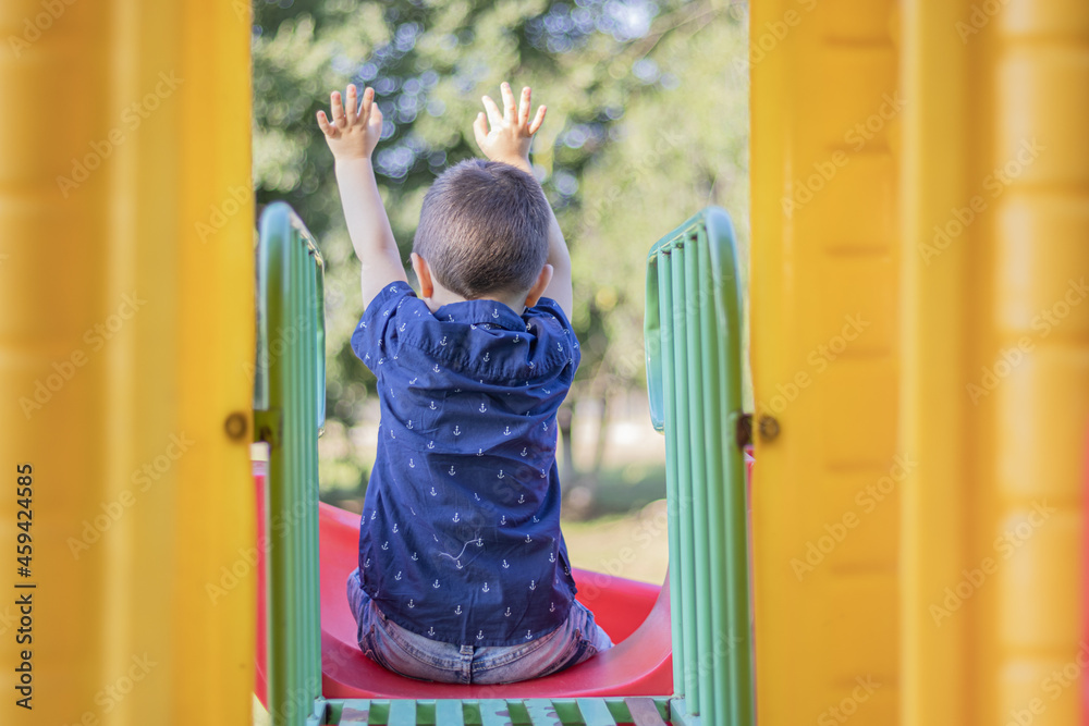 A 3 year old boy sliding on a red slide in an outdoor playground ...