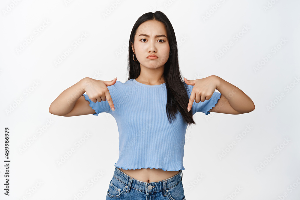 Suspicious young asian woman pointing fingers down, looking with doubt ...