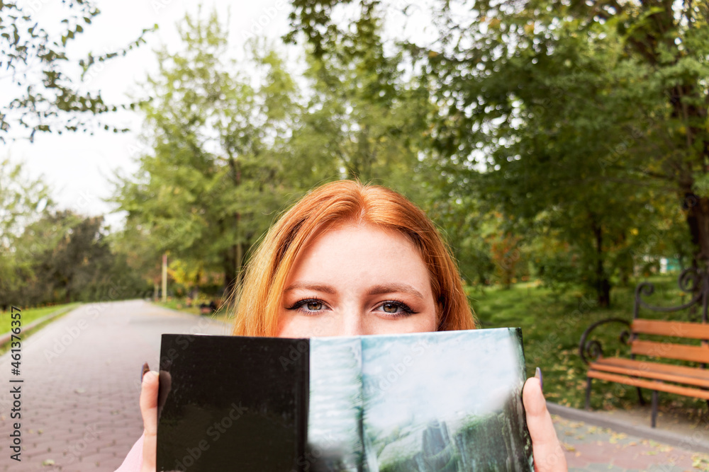 Young woman peeking out from behind an open book on a walk in the park ...