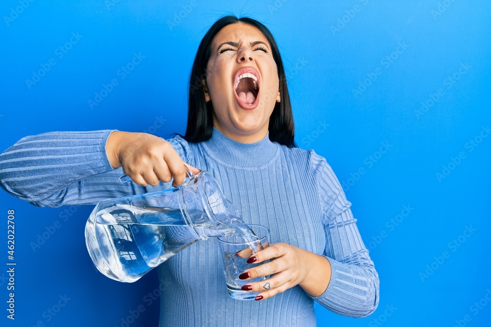 Beautiful brunette woman pouring water in glass angry and mad screaming ...