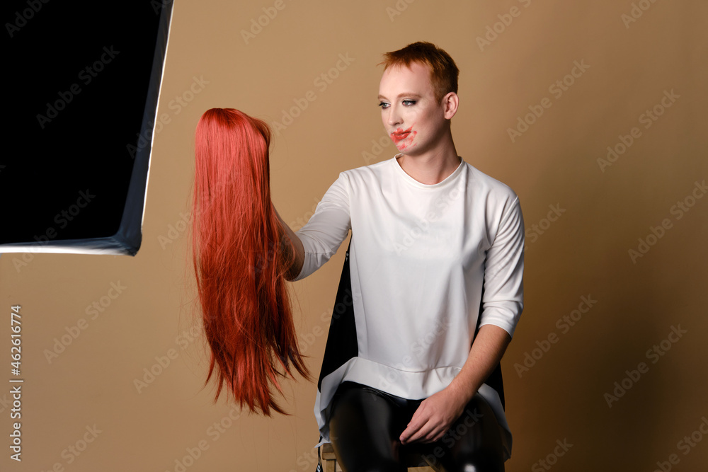 Man with makeup and smeared red lipstick, a wig in his hand. Portrait ...