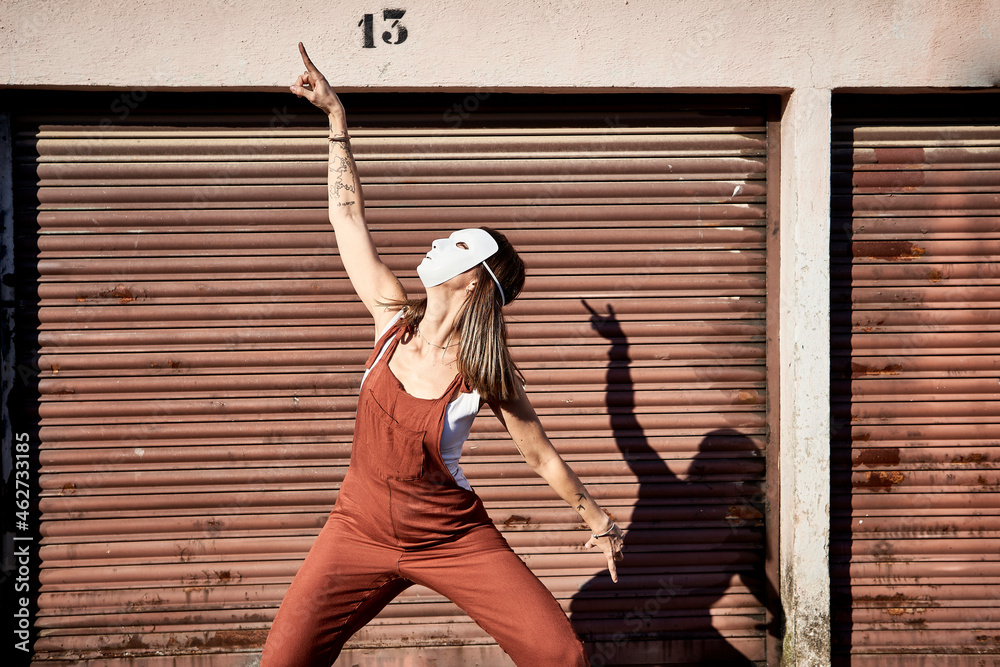 Woman wearing white mask dancing against closed shutter during sunny ...