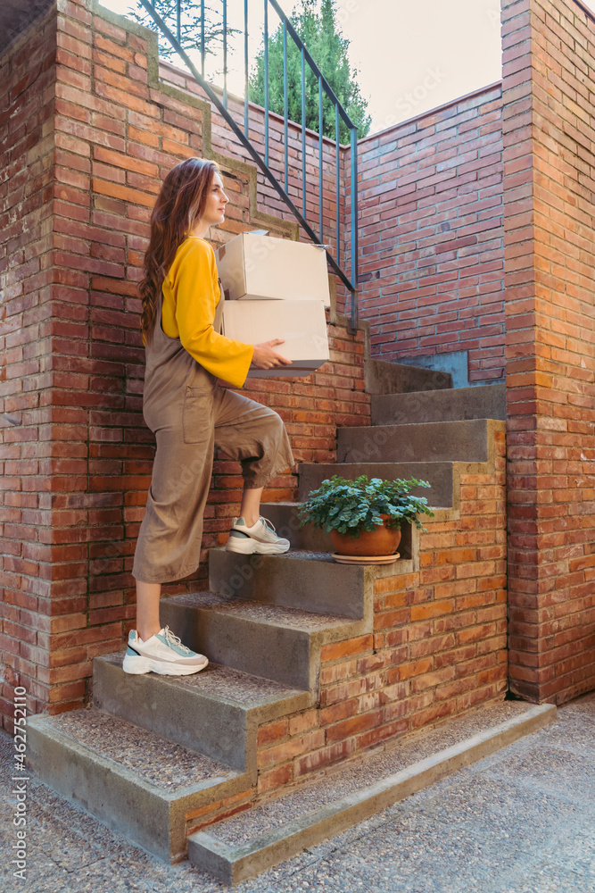 Young woman carrying white boxes while moving up on steps by brick wall ...