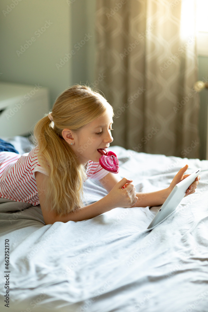 Blond Girl Lying On Bed Licking Lollipop While Using Digital Tablet