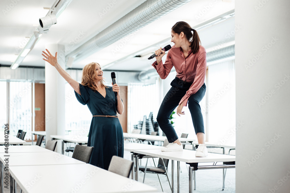 Happy female colleagues singing on microphone at conference table while ...