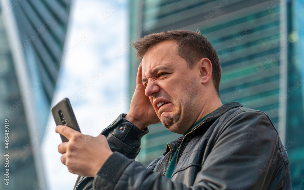 Sad millennial guy looks at phone screen in surprise and stands outside ...