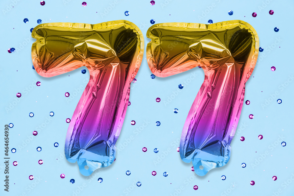 Rainbow foil balloon number, digit seventy seven on a blue background ...